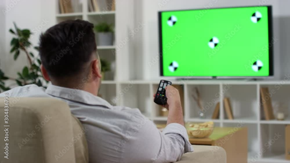 Back view of man walking into living room, sitting on sofa and using ...
