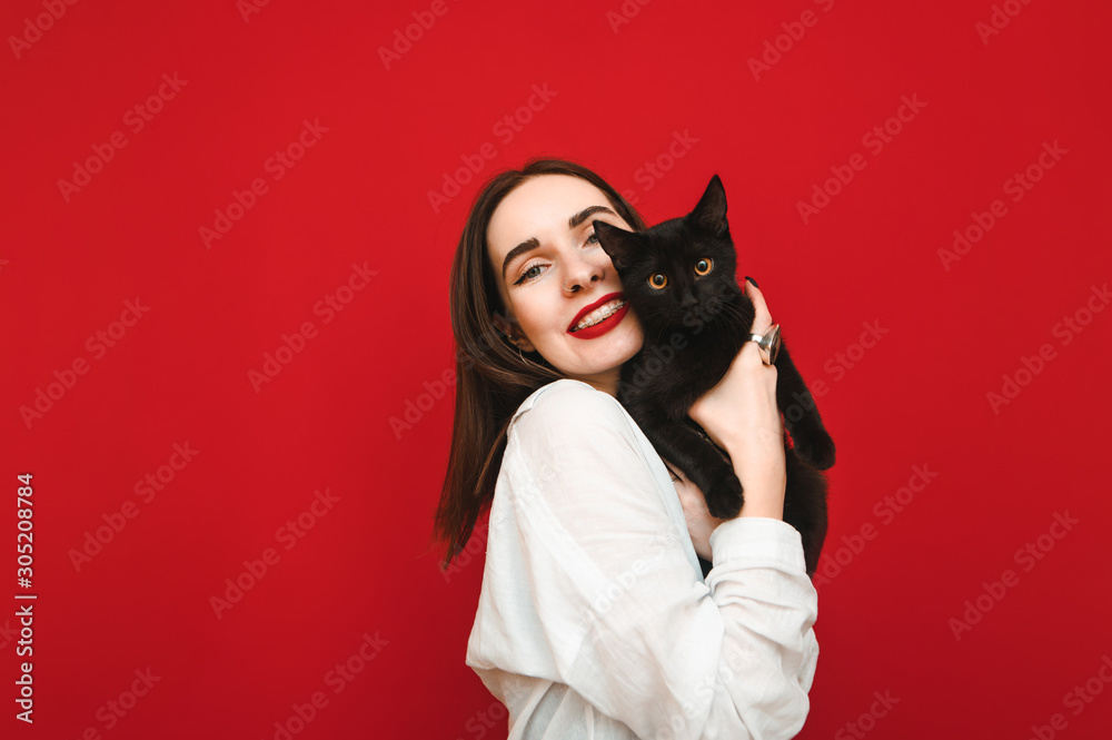 Portrait of a joyful girl hugging with a cute black cat on a red ...