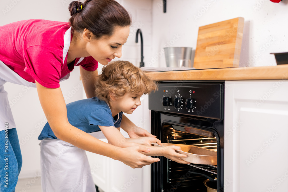 Little boy putting tray into open oven and his mom helping him while ...