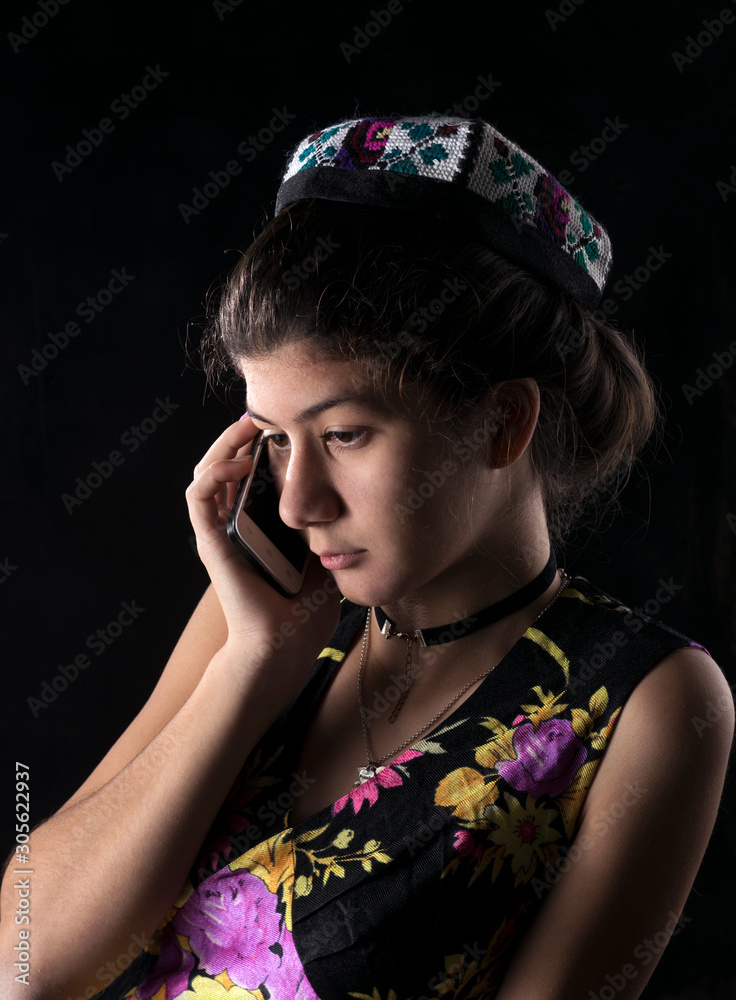 Beautiful girl in a national Central Asian skullcap and national dress talking on a smartphone ...
