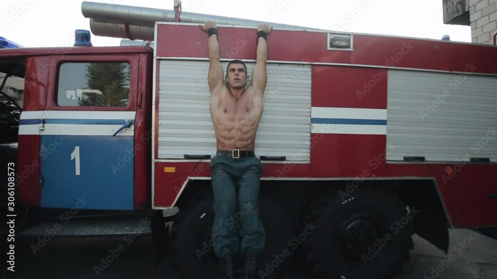 Muscular young man holding to the roof of a fire truck, raising his legs, training abdominal ...