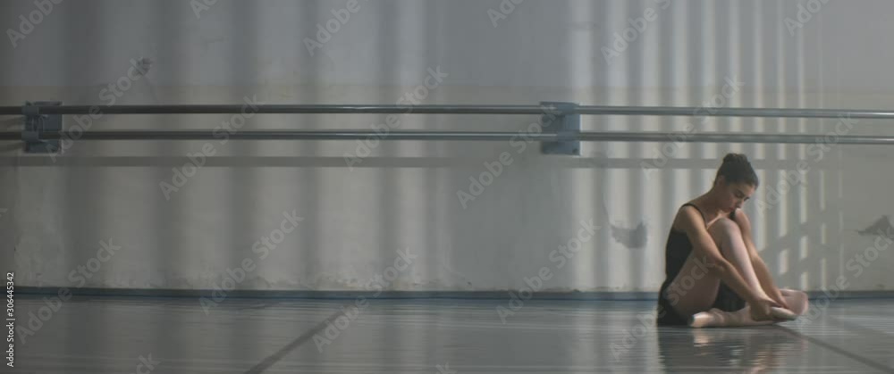 Pretty young caucasian ballerina is sitting on floor in dance hall ...