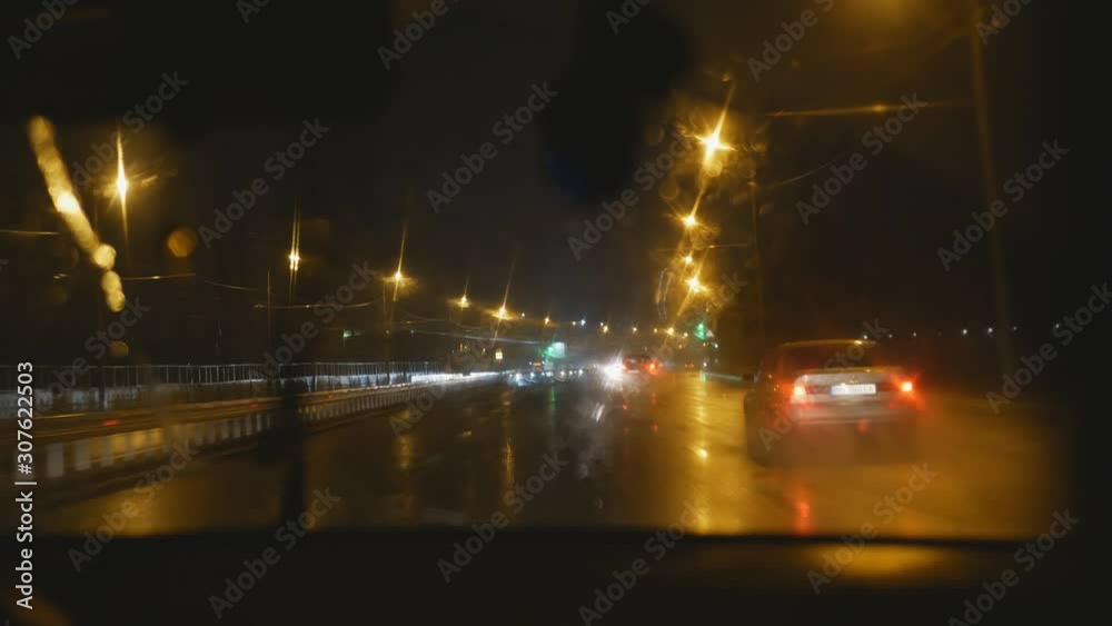Night city through the wet window of a moving car with a wiper.Rainy ...