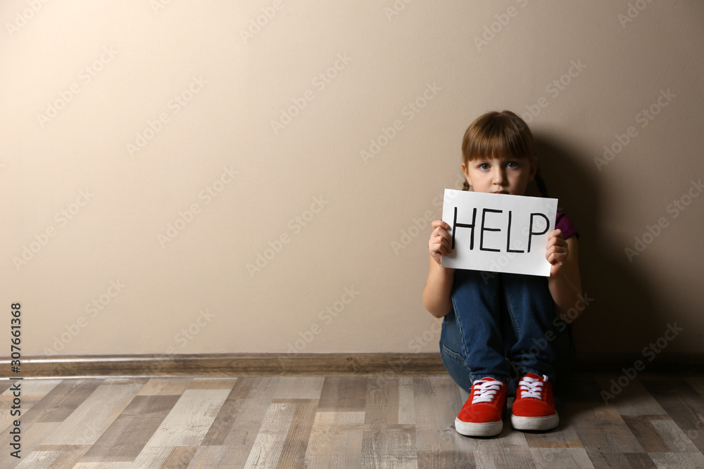 Sad little girl with sign HELP sitting on floor indoors, space for text ...