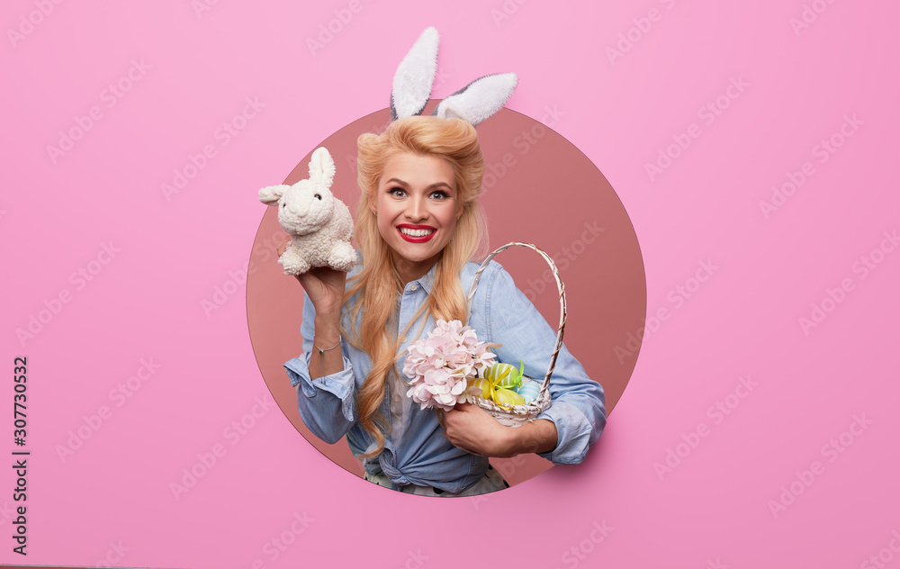 Happy Easter. Young woman with bunny ears and basket full of Easter eggs on pink background ...