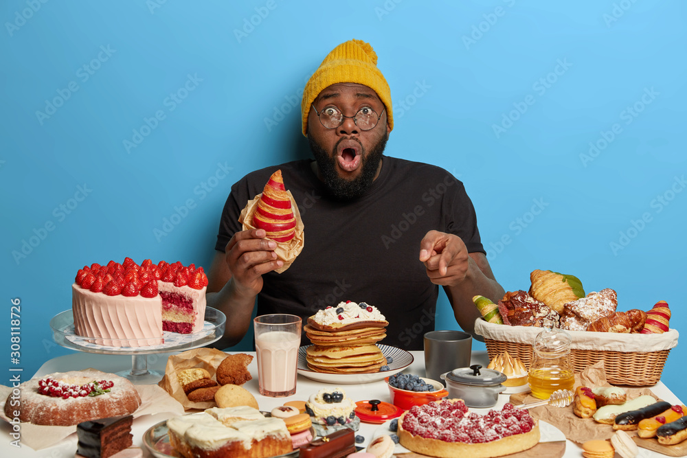 Stupefied black man eats tasty croissant, points at table full of sweet ...