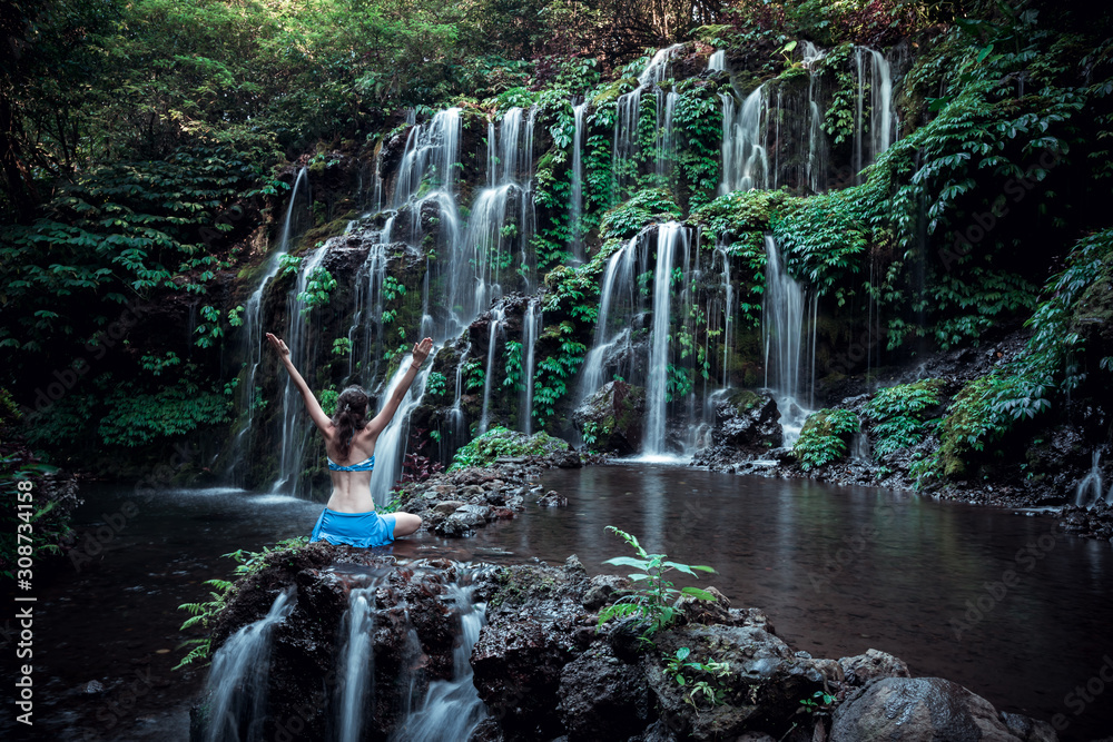 Excited Caucasian woman raising arms in front of waterfall. View from ...