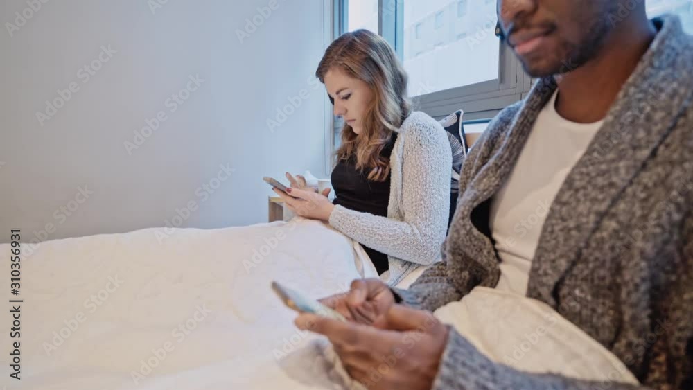 Multiracial couple sitting in their bed typing in their cells and ...