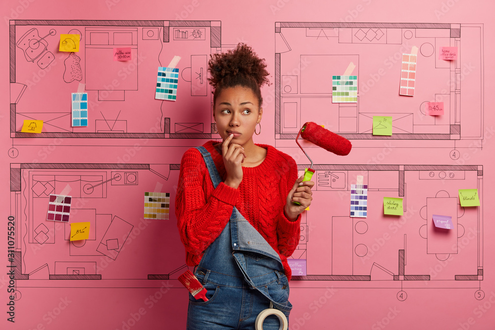 Pensive female homemaker applies red paint with roller, wears sweater ...