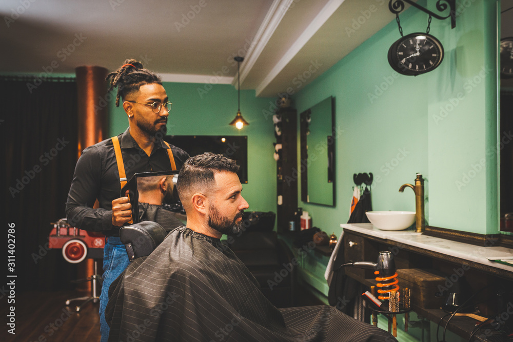 un barbero afroamericano de estilo hipster enseña a su cliente blanco ...