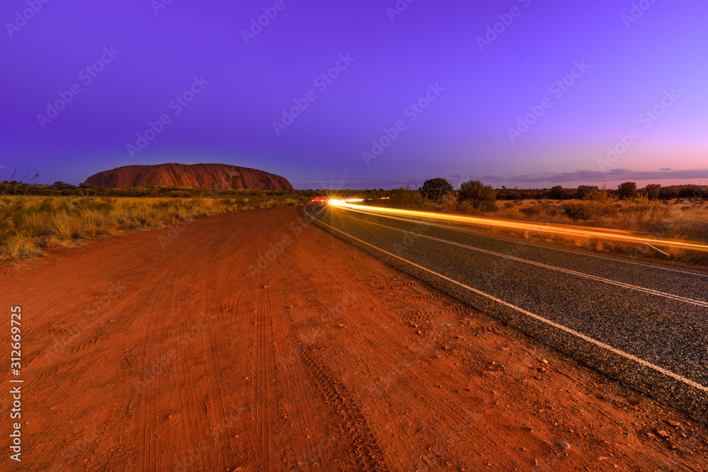 Car light trails along the road to Uluru Ayers Rock at night. The huge ...