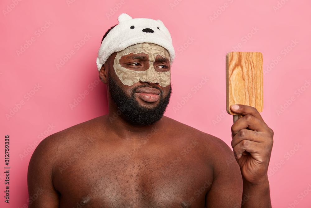 Headshot of puzzled dark skinned man applies facial clay mask, looks in ...