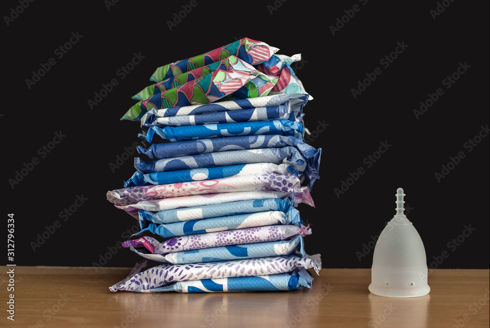 A silicone menstrual cup next to a stack of sanitary towels and tampons ...