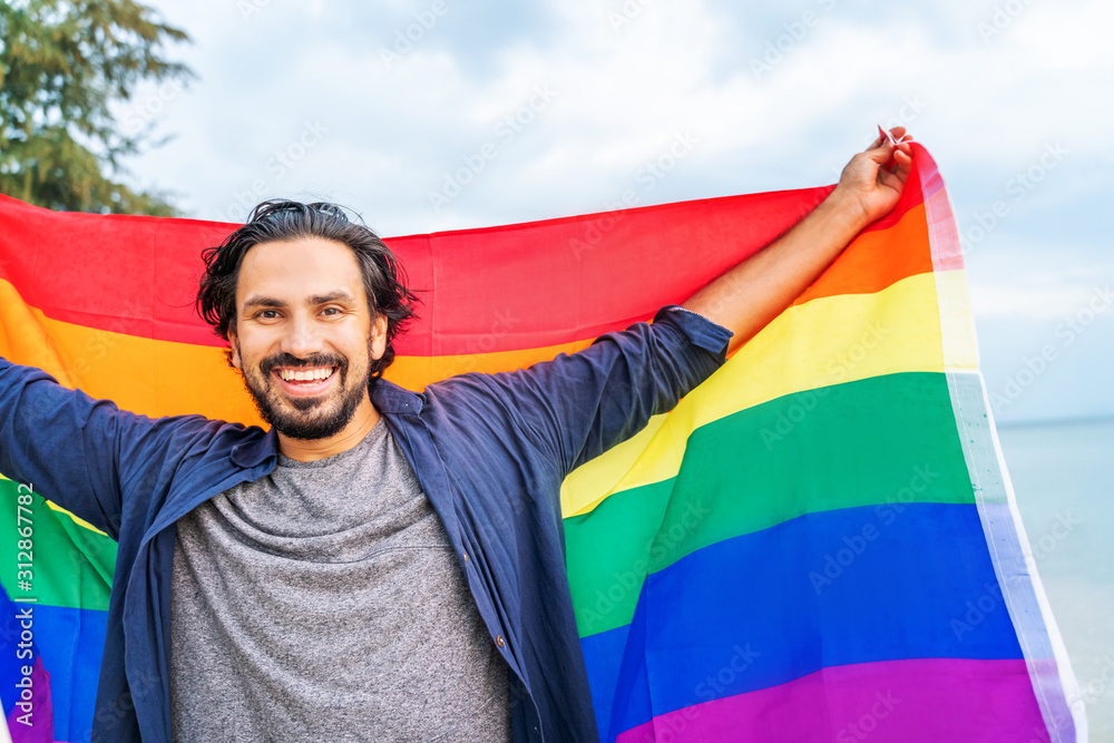 Cheerful guy with a rainbow flag on the beach. Young man holding a ...