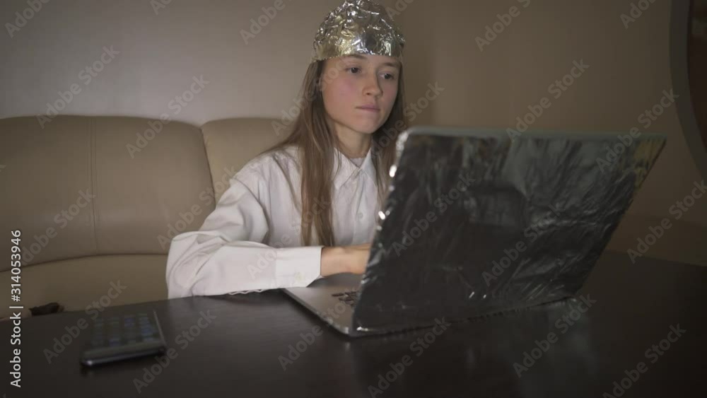 Young woman in Tin Foil Hat typing message on keyboard, laptop with foil. Foil hat shields her ...