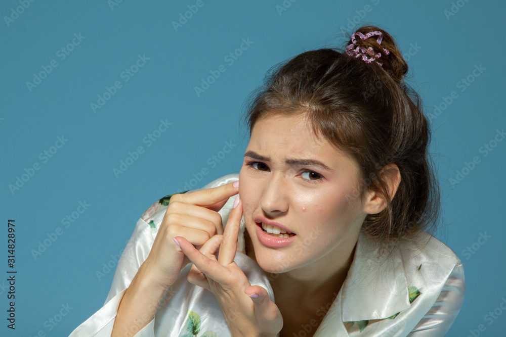 Teen girl with problem skin look at pimple, blue background. Morning ...
