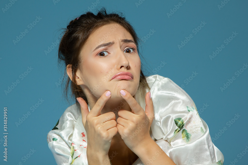 Teen girl with problem skin look at pimple, blue background. Morning ...