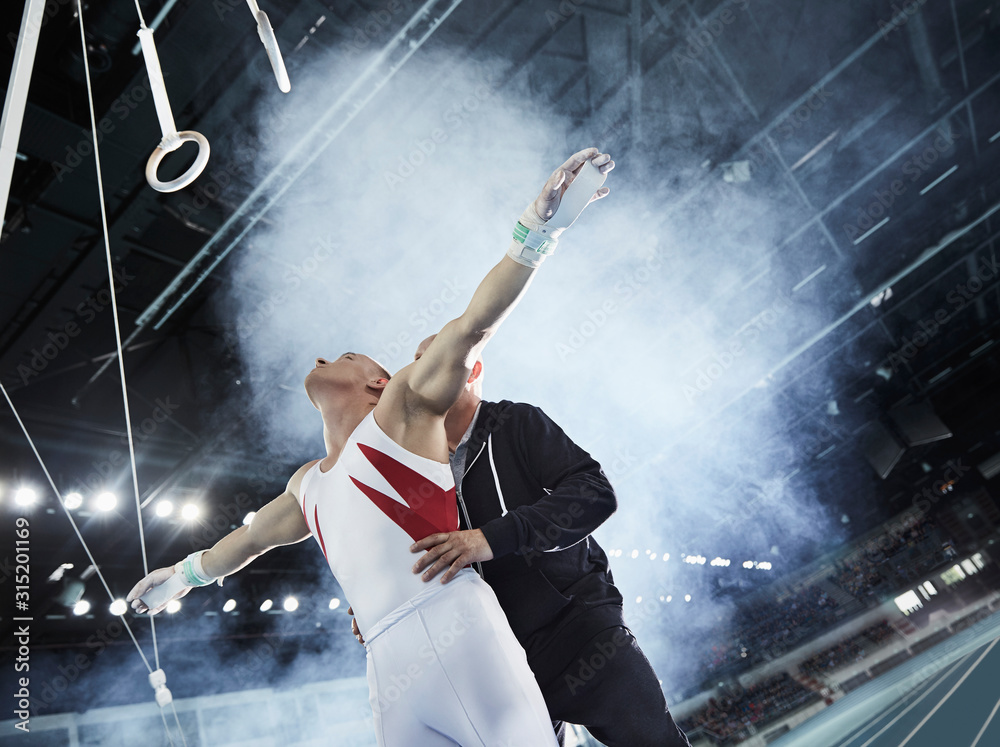 Coach lifting male gymnast below gymnastics rings in arena Stock 写真 ...