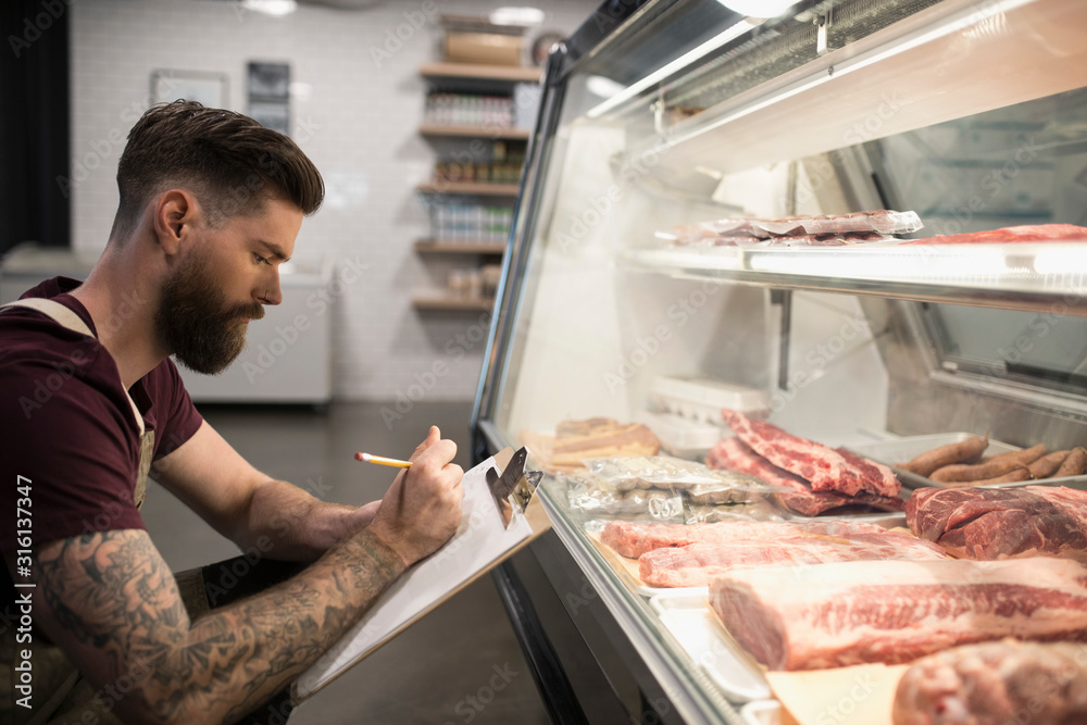 Male butcher with clipboard checking raw red meat inventory at display ...