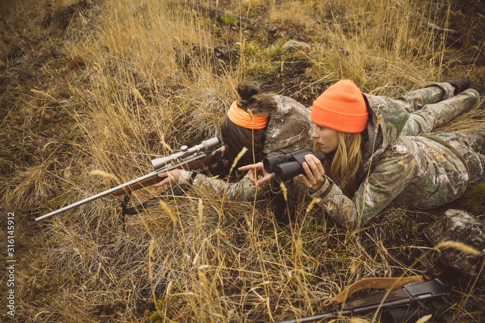 Mother and daughter hunters laying, using binoculars and hunting rifle ...
