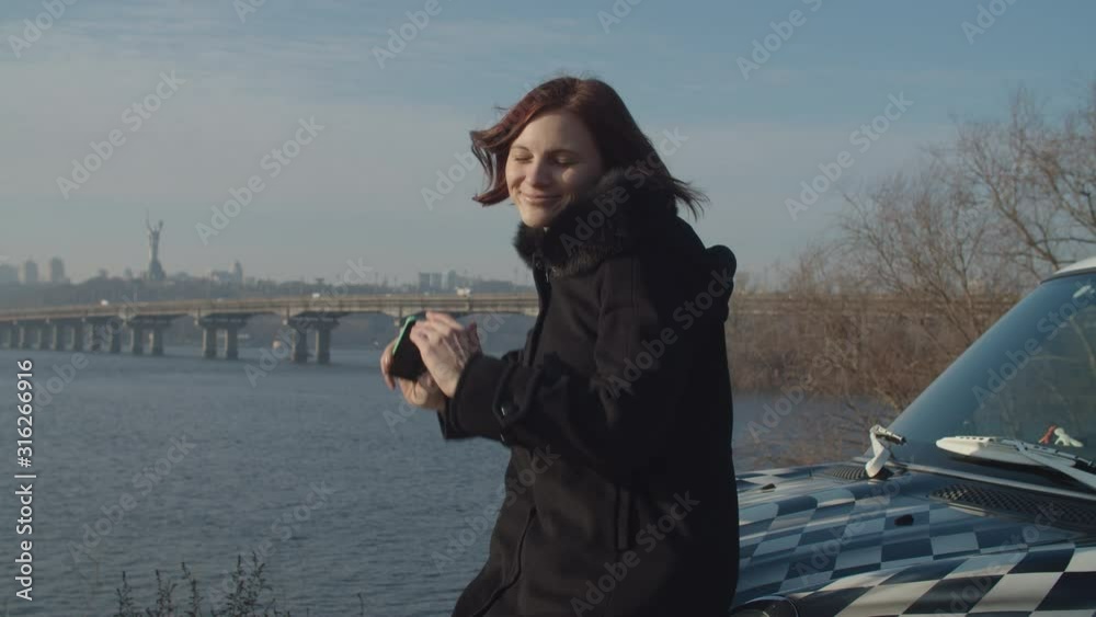 Laughing female 30s adult in black coat on car hood dancing with ...