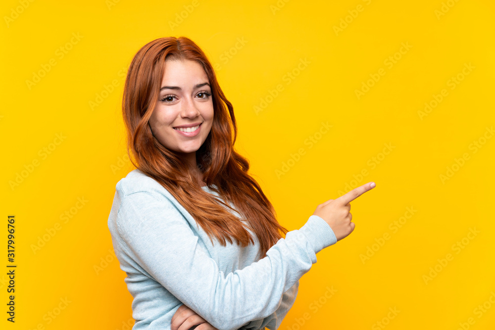 Teenager redhead girl over isolated yellow background pointing finger ...