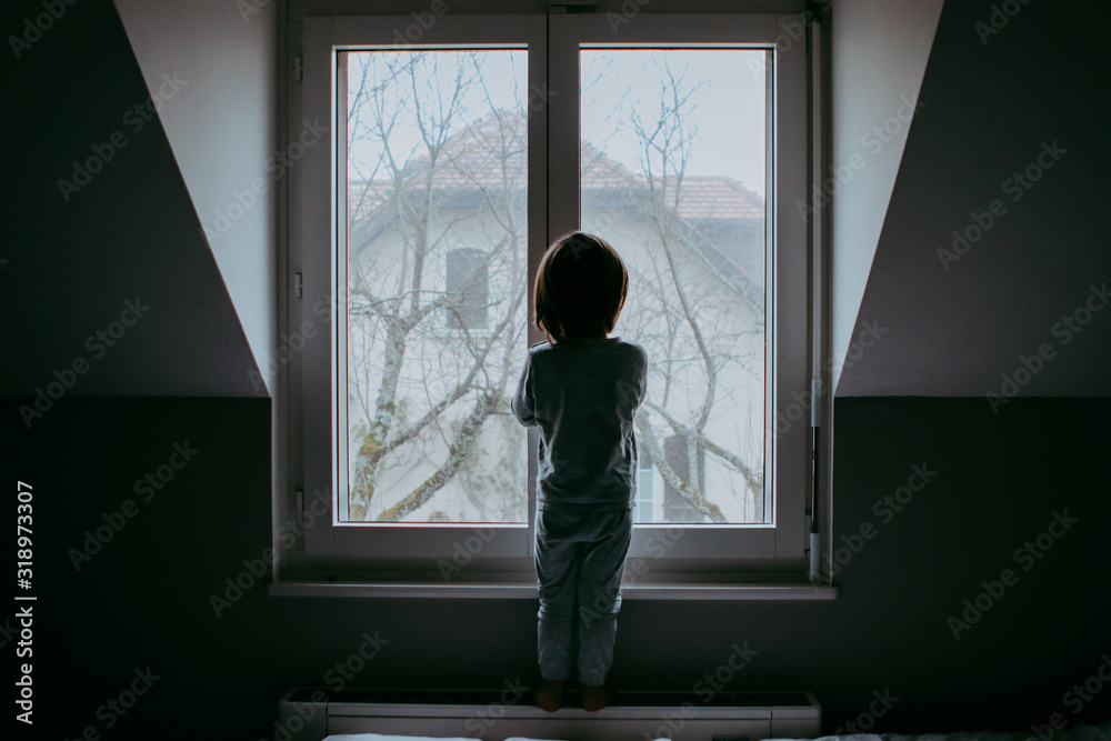 Back view of little child standing at closed window in dark room ...