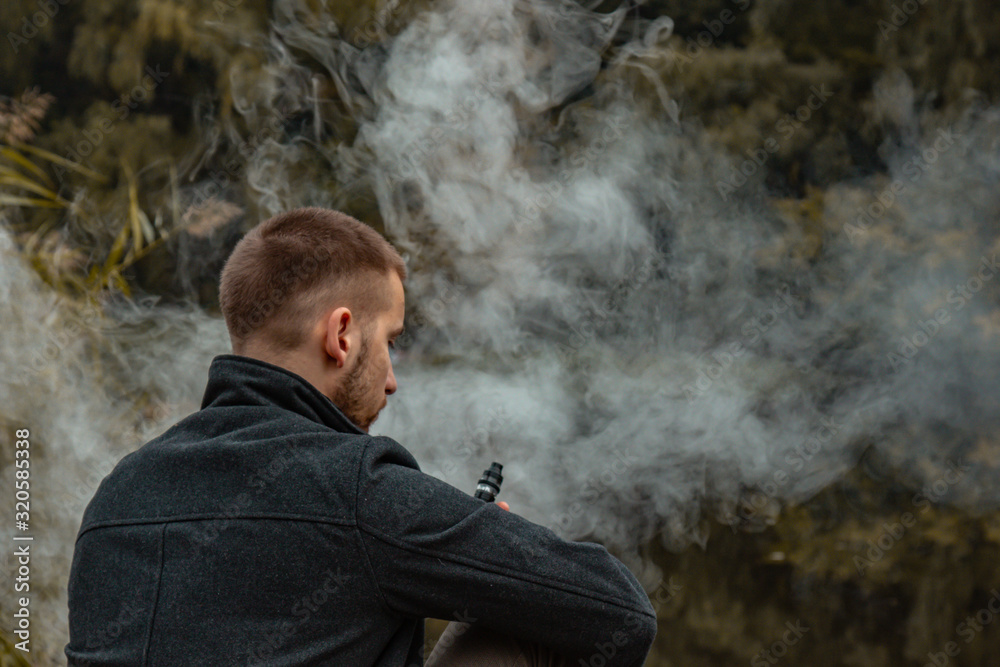 A guy with a beard sits on the lake and smokes an electronic cigarette ...