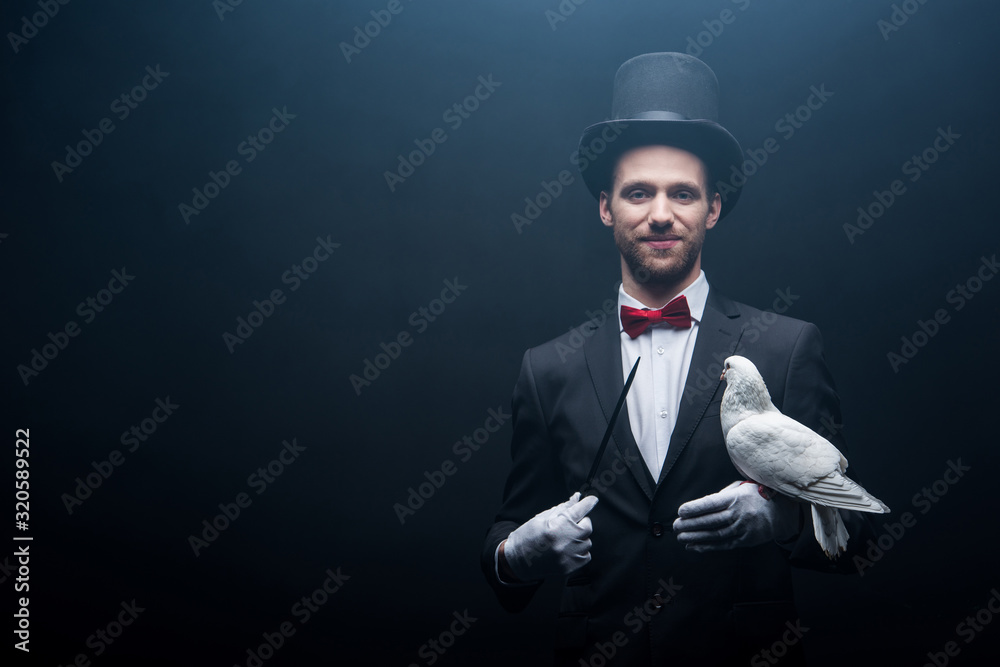 smiling magician in hat showing trick with dove and wand in dark room ...