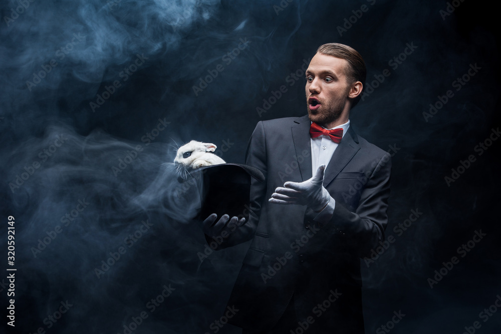 surprised magician in suit showing trick with white rabbit in hat, dark ...