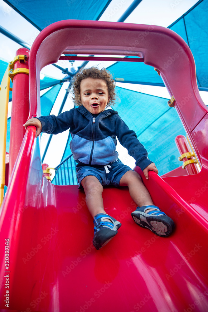 Happy cute young diverse boy with enjoying a ride down playground slide ...