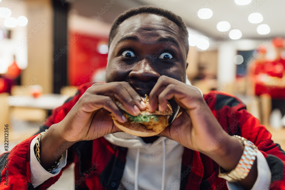 A portrait of young african man eating a burger in street food cafe ...