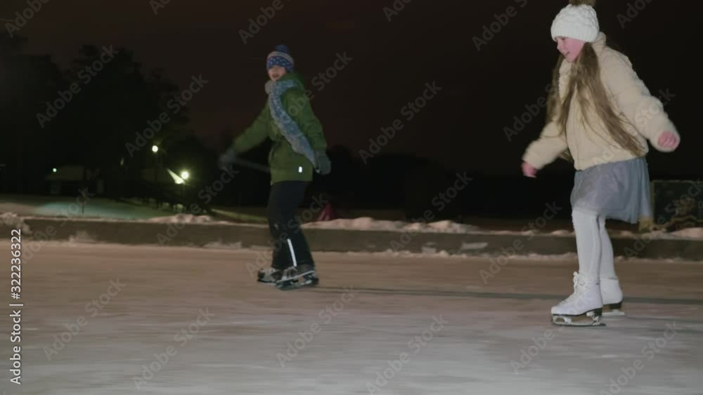 Happy boy and girl teenagers fast skating on ice rink at winter dark ...