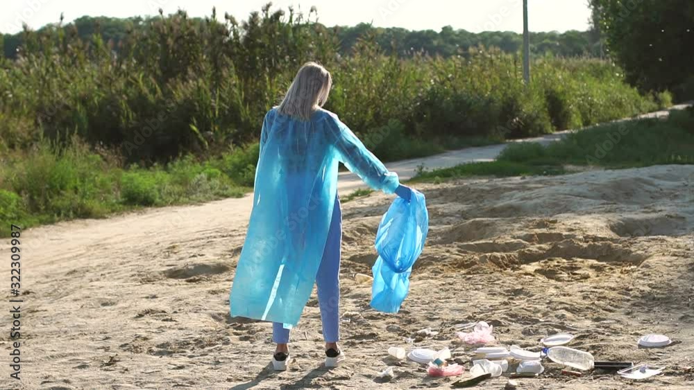 A volunteer girl in a Bathrobe and gloves collects garbage on the beach ...