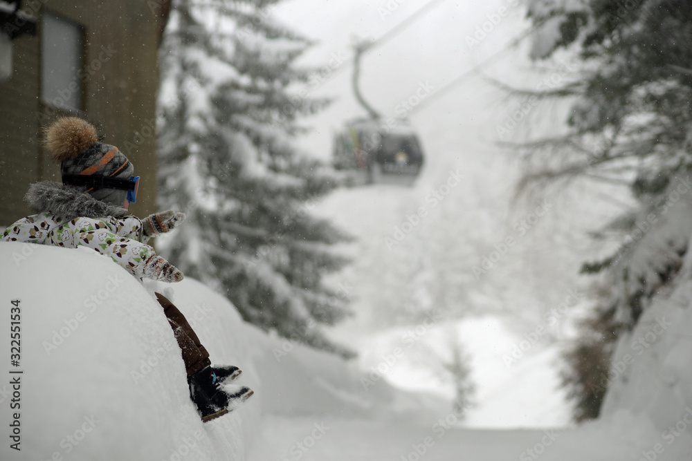 Small child in ski mask sitting on big snowbank under snowfall ...