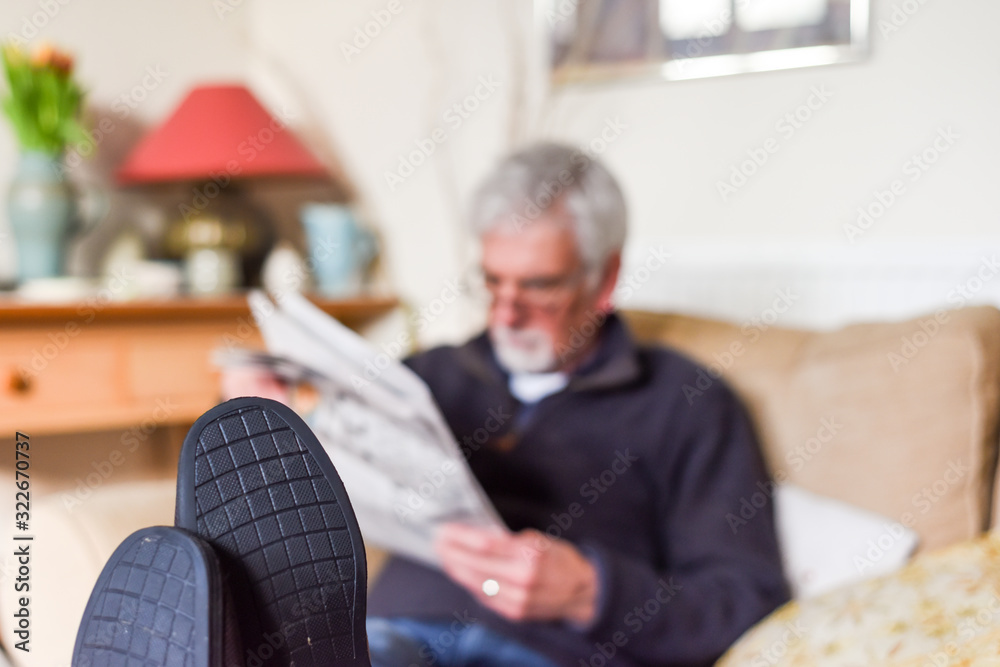 Mature retired man with feet up sitting on a sofa reading a newspaper ...