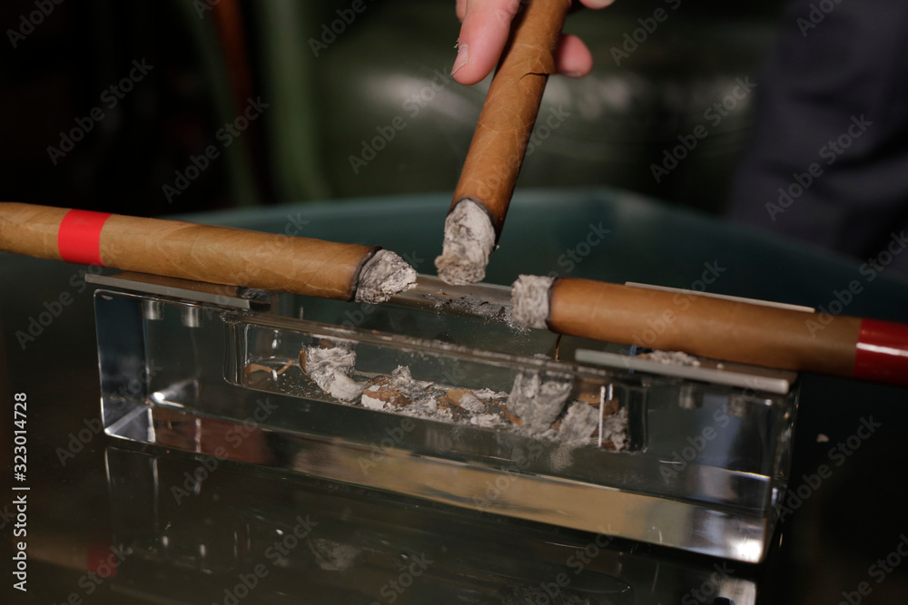 two smoking cigars lie in a transparent ashtray, a man brings a third ...