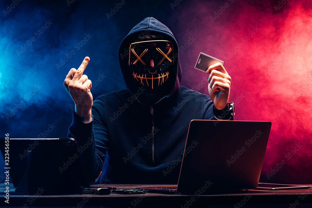 young impudent masked hacker isolated over smoky space, sit showing middle finger in the meaning ...