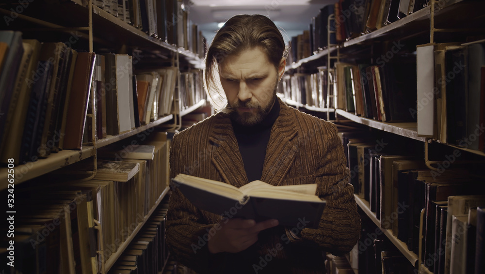 Unshaven male captured with reading standing between rows of bookshelf ...