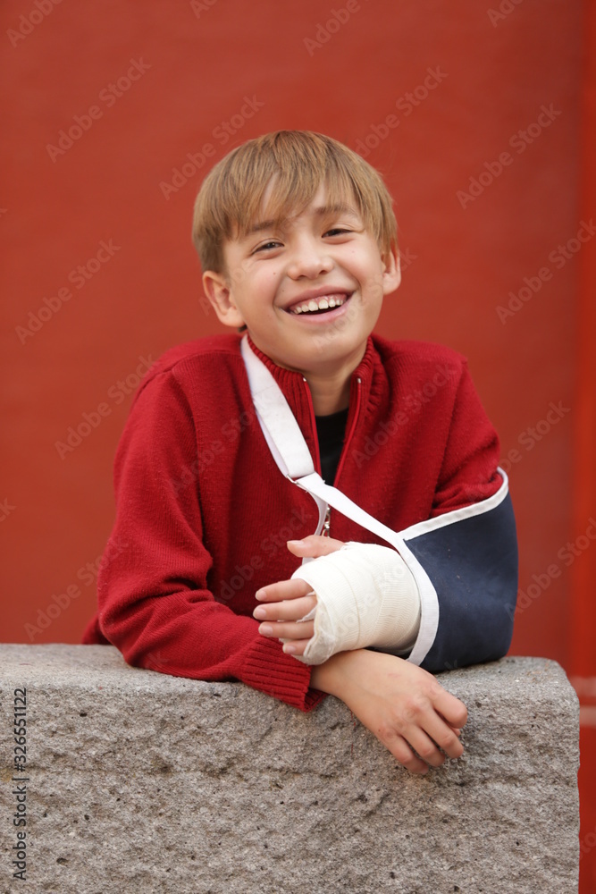 Smiling blond boy with bandaged arm with plaster and sling. Left arm ...