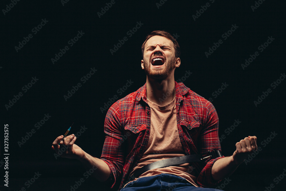 amazed caucasian junkie man sit shouting isolated over black background ...