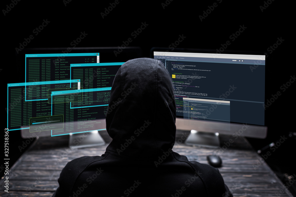 back view of hooded hacker sitting near computer monitors with data on ...