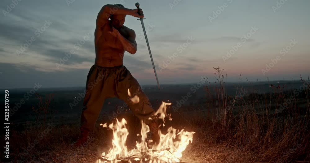 Powerful muscular shirtless man wielding a sword over the flames of a ...