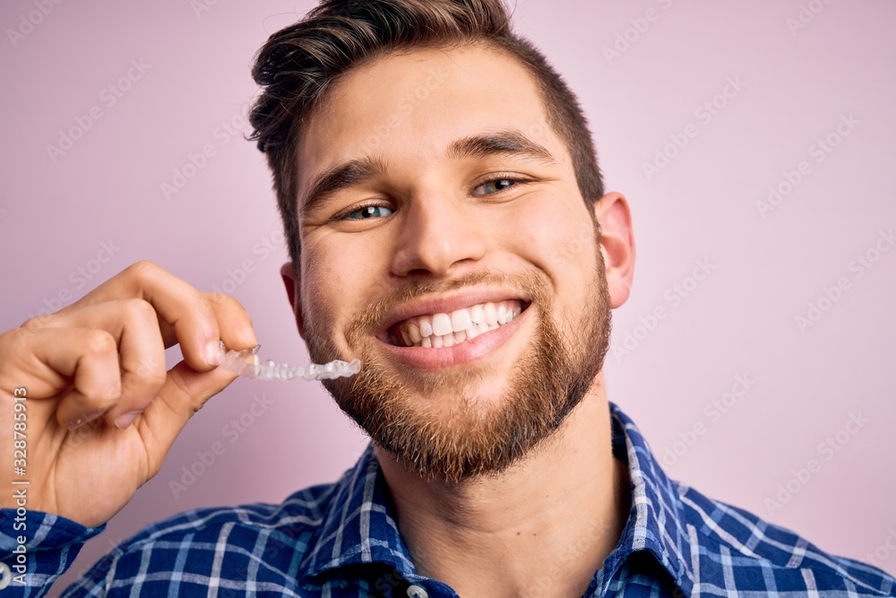 Young blond man with beard and blue eyes holding dental aligner over ...