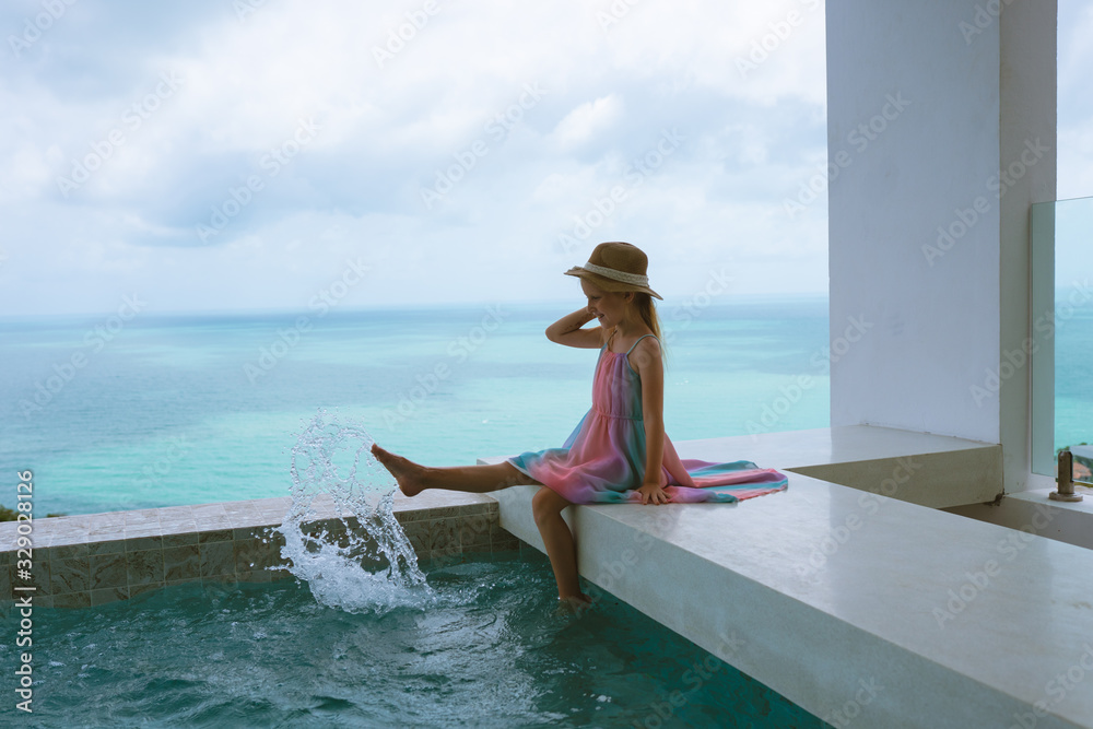 Little and beautiful girl with red hair and freckles sits by the pool ...