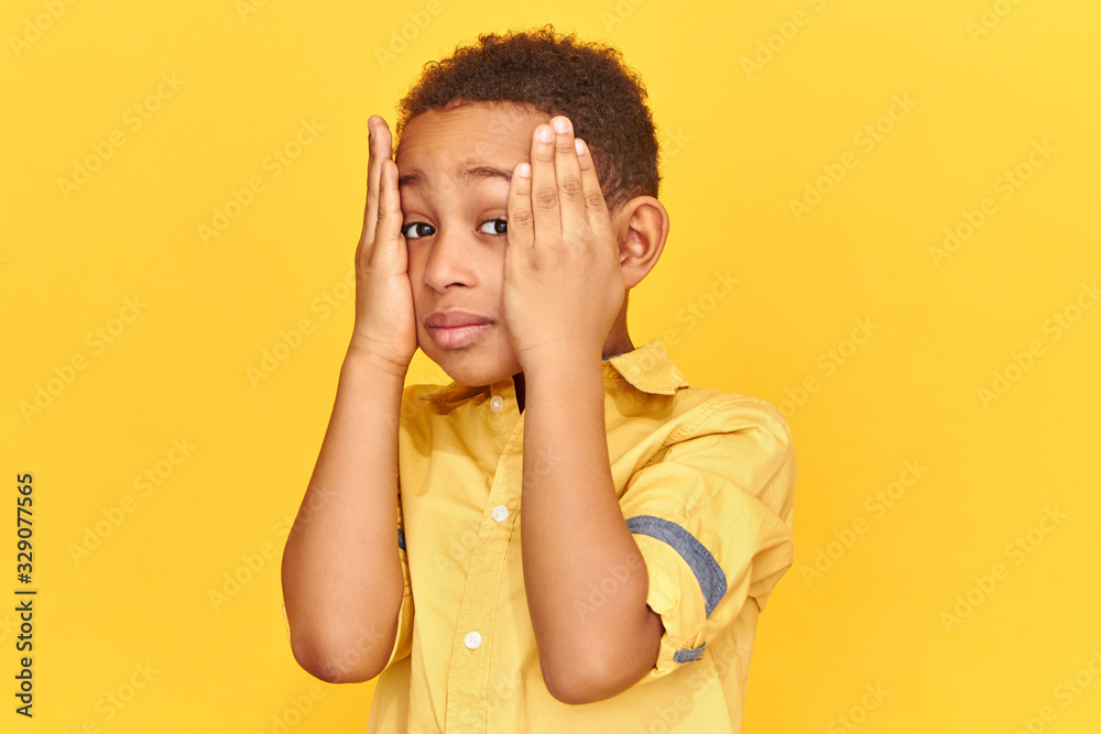 Emotional dark skinned child keeping hands on his cheeks having upset ...