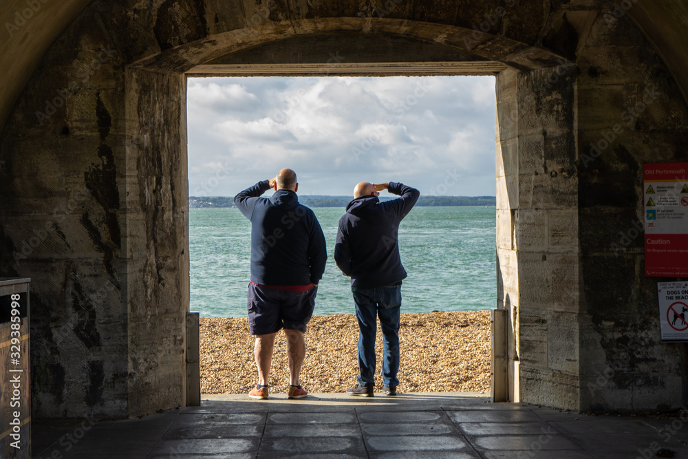 two men stood in an archway at the beach looking out to sea shading ...