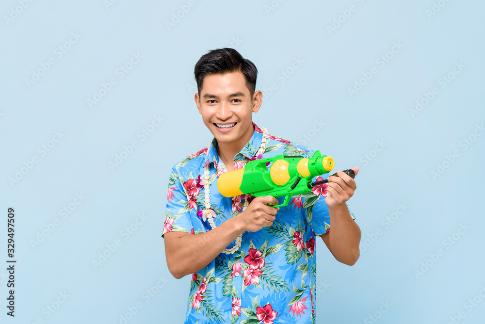 Smiling handsome Asian man playing with water gun isolated on studio ...