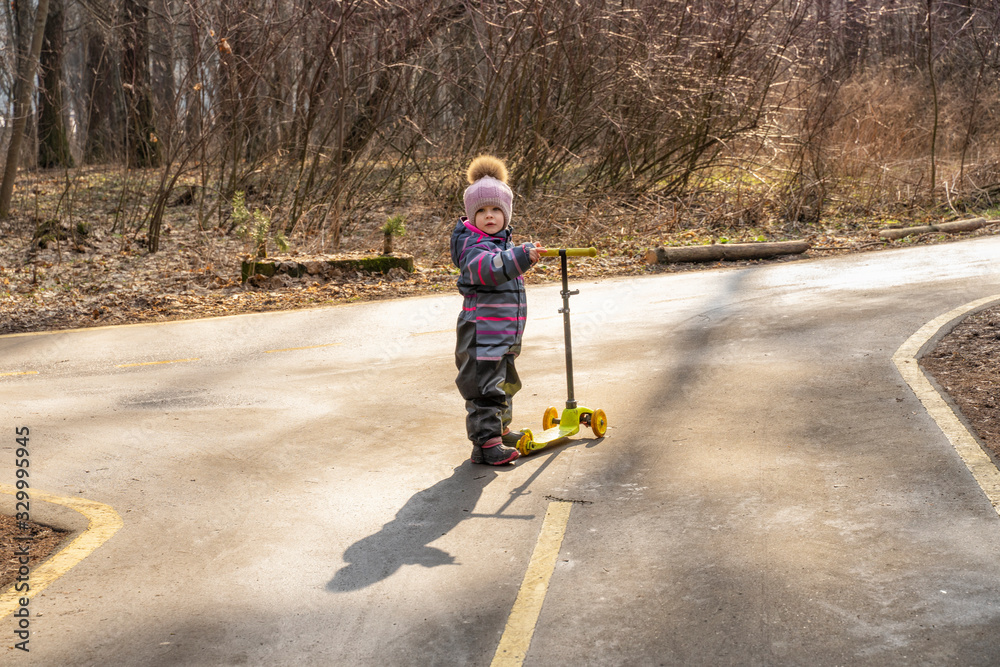 child with a scooter on an asphalt road in a spring park in a hat with ...