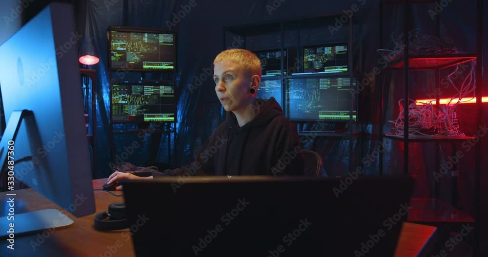 Female Caucasian hacker working on computer in front of big screen and typing on keyboard in ...